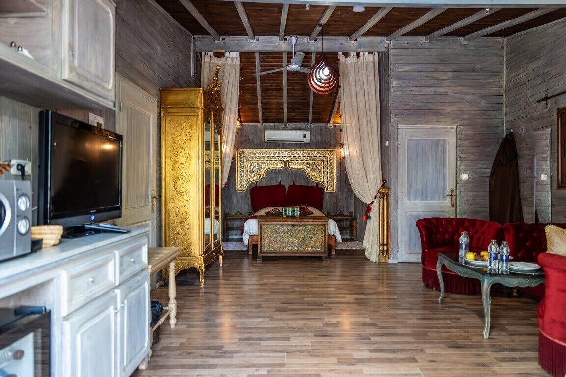 Um quarto com piso de madeira, cama com cabeceira vermelha, sofá vermelho, televisão sobre um armário e ventilador de teto.