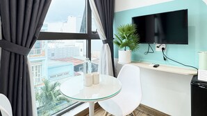 Quarto de hotel moderno com uma janela ampla, uma mesa de vidro redonda com duas cadeiras, uma TV de tela plana na parede e um pequeno vaso de planta numa prateleira.