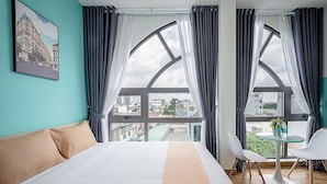 Quarto de hotel com uma cama grande, duas janelas com cortinas, uma varanda com mesa e cadeiras, e vista para a cidade.