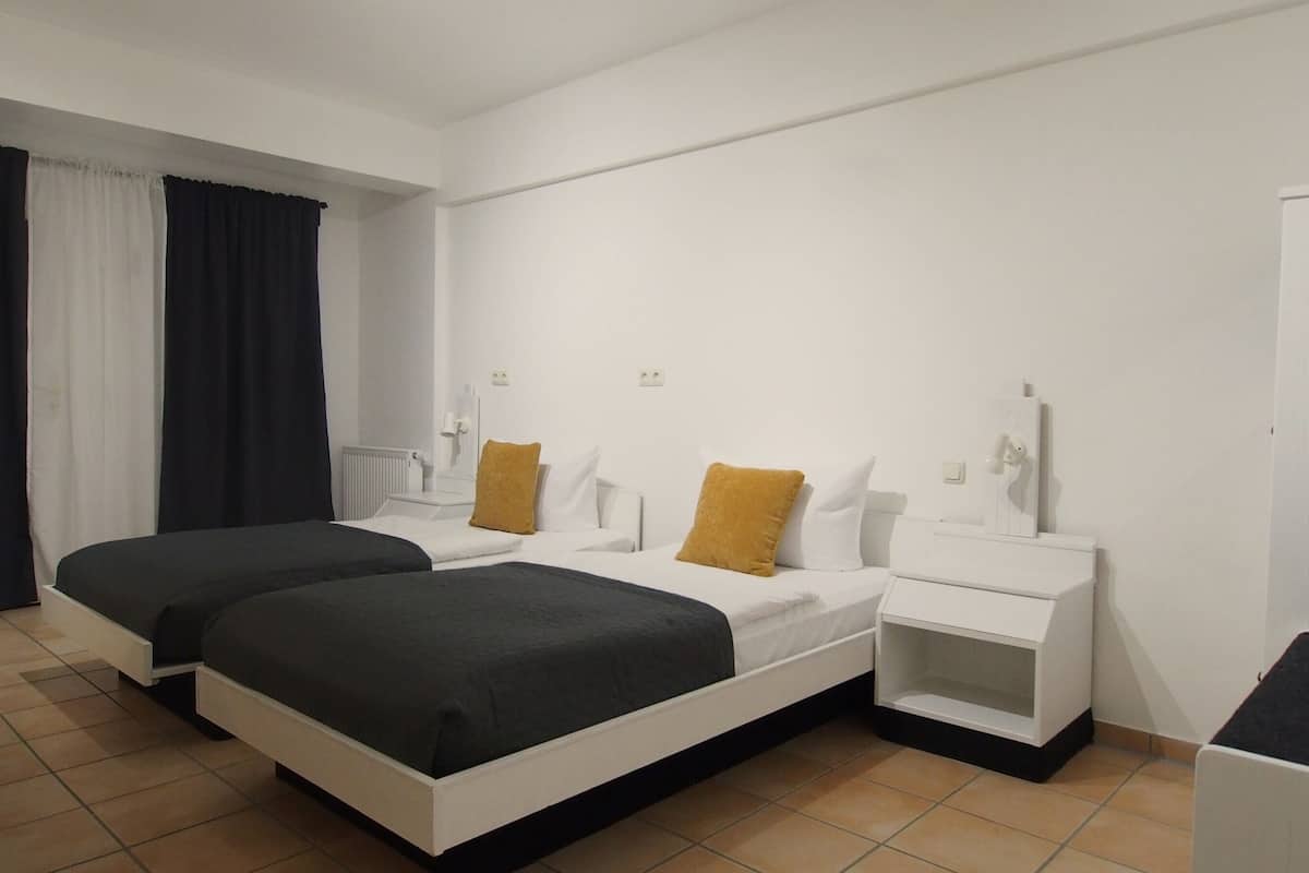 Quarto de hotel com duas camas de solteiro, mesas de cabeceira brancas, uma pequena mesa e uma janela com cortinas preto e branco.