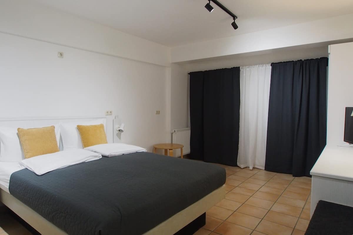 Quarto de hotel com uma cama, mesas de cabeceira brancas, uma pequena mesa e uma televisão.