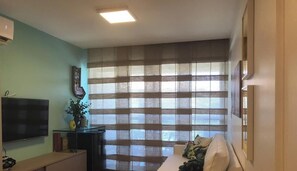 Uma sala de estar com televisão, sofá e uma grande janela com persianas.