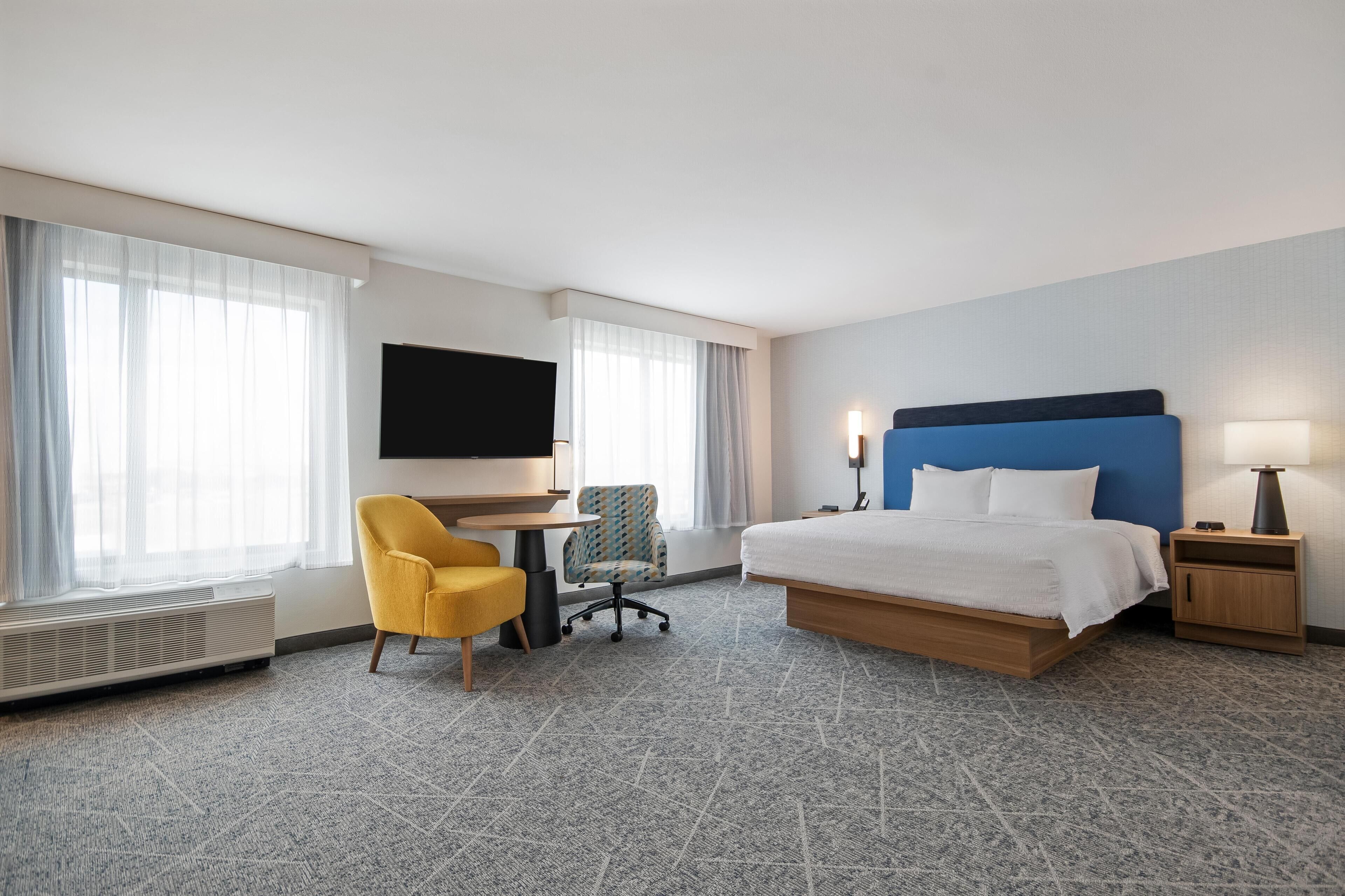Quarto de hotel com cama, escrivaninha, cadeira, televisão e janela com cortinas.