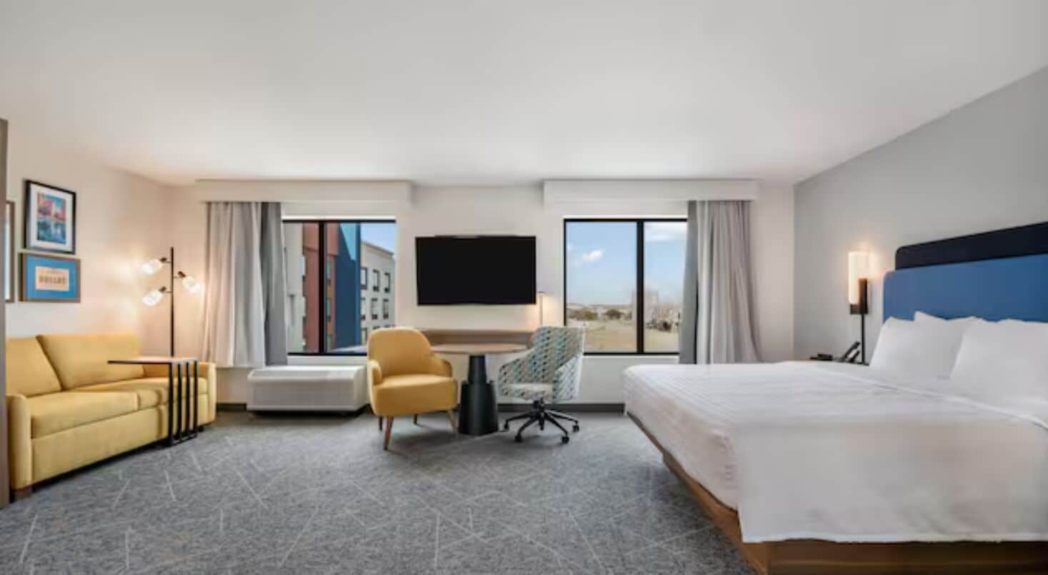Quarto de hotel com cama, sofá, escrivaninha, cadeira, televisão e janelas amplas.