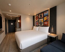 Quarto de hotel com uma cama grande, uma mesa de cabeceira, uma cadeira, ar condicionado fixado na parede, uma porta e uma parede com obras de arte emolduradas.