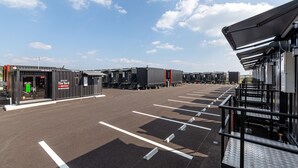 Um parque de estacionamento com fileiras de contentores de transporte e um edifício com a inscrição 'The Yard', com uma placa a indicar que é um hotel.