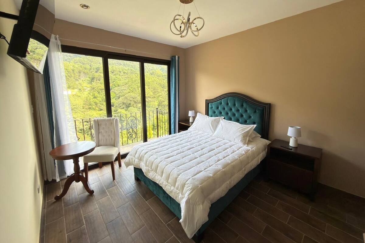 Quarto com uma cama grande, uma cadeira, uma mesinha e vista para o verde lá fora.