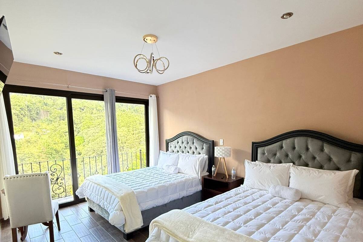Quarto com duas camas, uma cadeira, uma mesinha de cabeceira e vista para área verde através de grandes janelas.