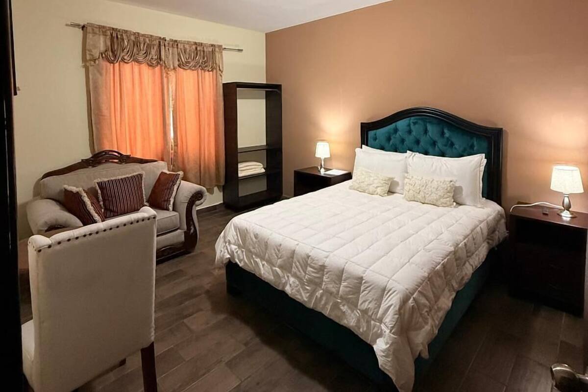 Quarto de hotel com cama, cadeira, mesa de cabeceira, abajur e espelho.