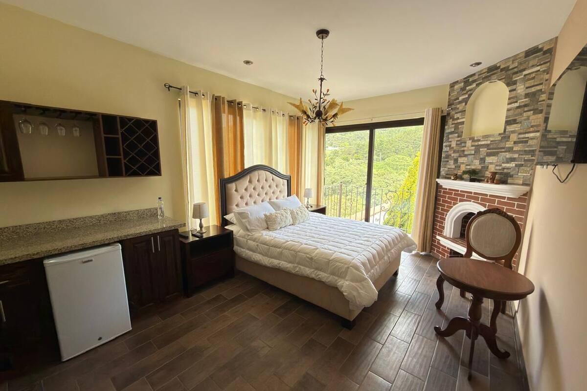 Quarto com cama, pequena mesa, cadeira, frigorífico embutido, lareira e uma janela grande com cortinas.