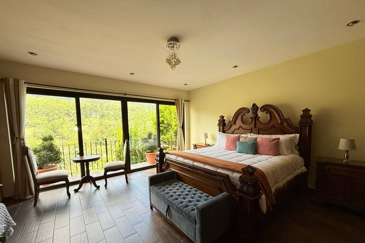 Quarto com uma cama grande, um banquinho, uma mesinha e vista para o verde através de janelas amplas.