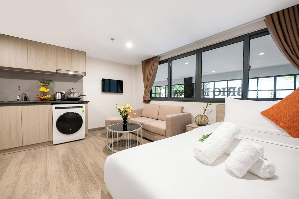 Quarto de hotel moderno com cama, sofá, pequena cozinha e janela com cortinas.