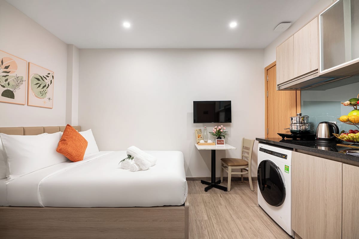 Quarto de hotel compacto com cama, uma pequena mesa com televisão, máquina de lavar e cozinha compacta.