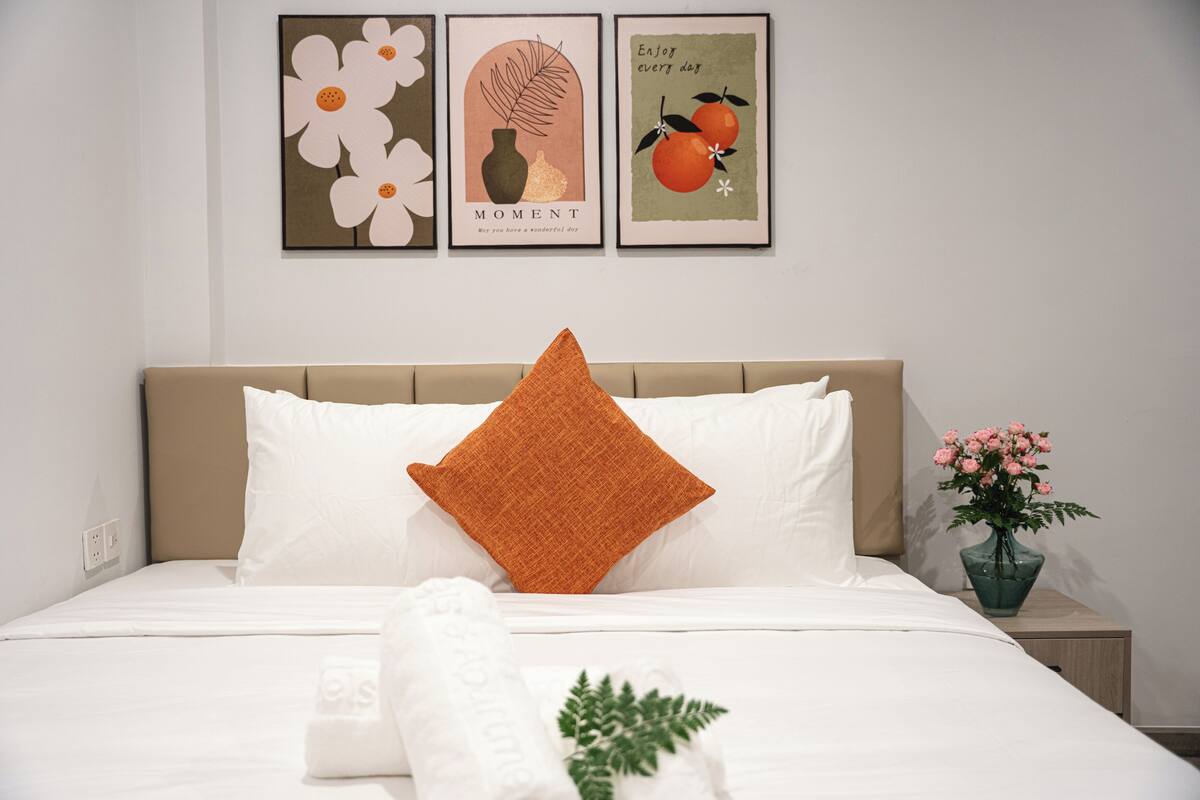 Uma cama bem arrumada com lençóis brancos, um único travesseiro laranja e um pequeno vaso com flores cor-de-rosa em uma mesa de cabeceira.