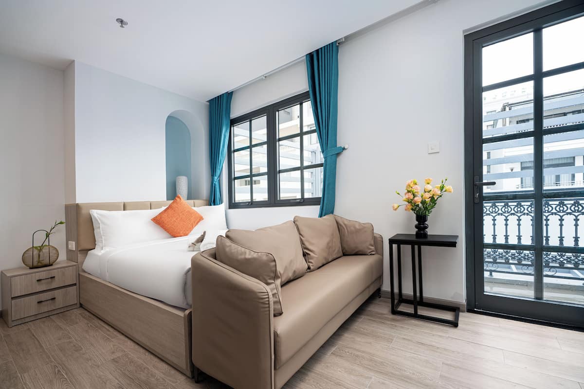 Quarto de hotel moderno com cama, sofá, mesa de cabeceira, pequena mesa com um vaso de flores e uma grande janela com vista para prédios.