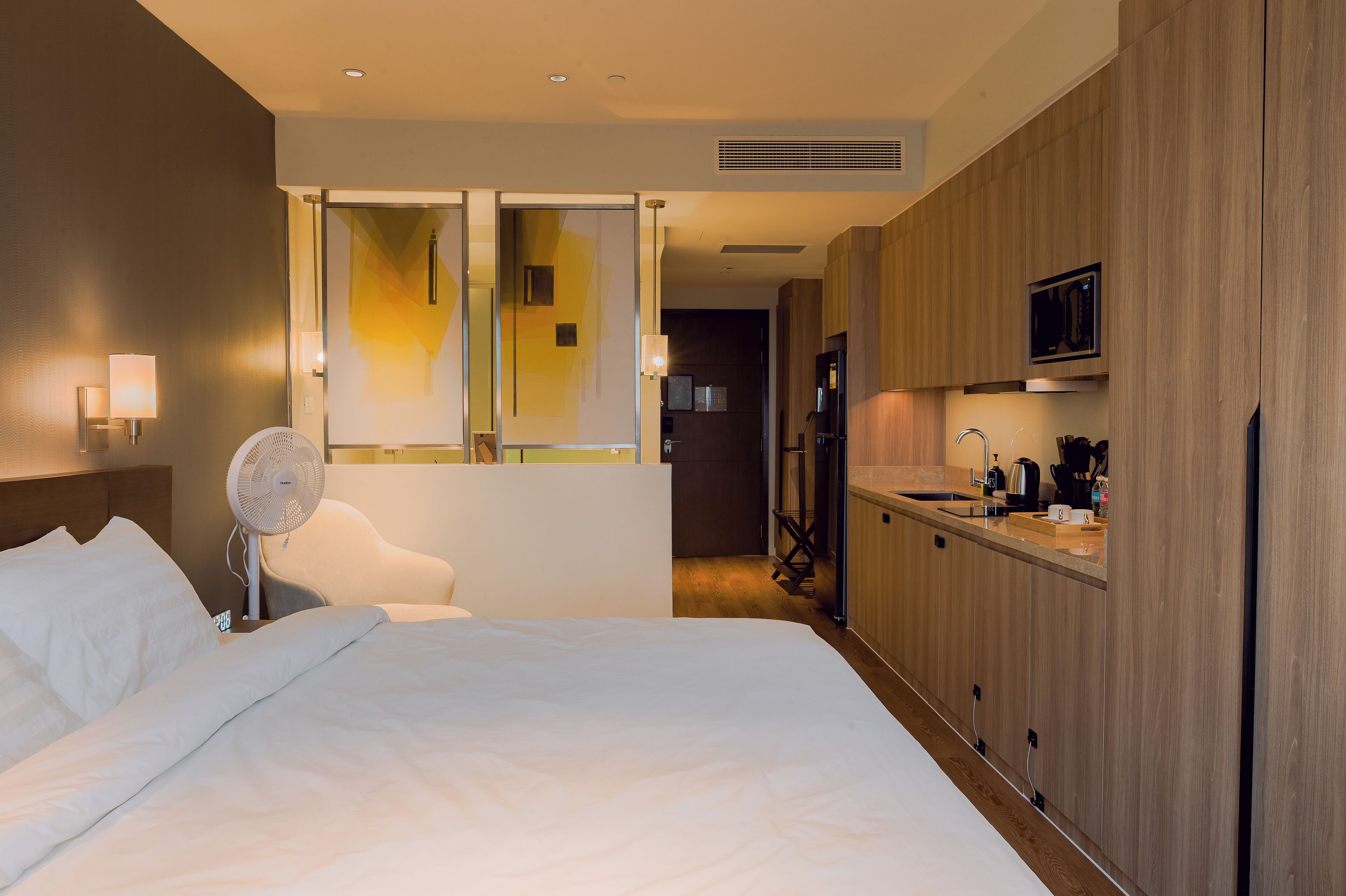 Quarto de hotel com cama, ventoinha na mesinha de cabeceira e uma cozinha compacta com micro-ondas.
