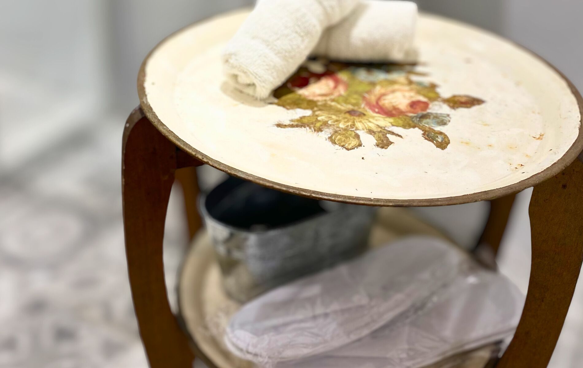 Uma pequena mesa redonda com um desenho floral no tampo, um tecido branco e um objeto preto por baixo.