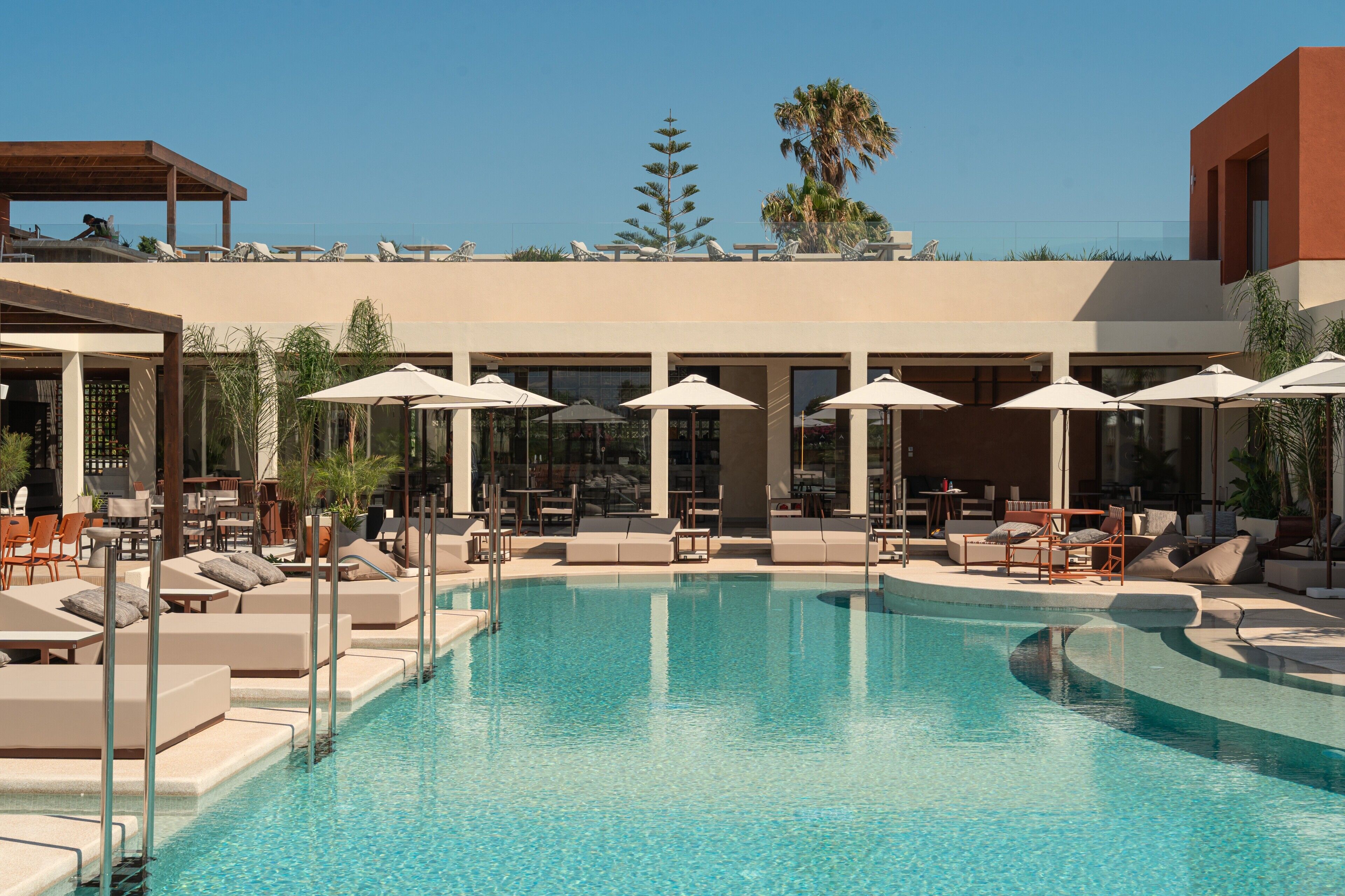 Área de piscina com espreguiçadeiras e guarda-sóis, cercada por um edifício com grandes janelas e uma palmeira ao fundo.