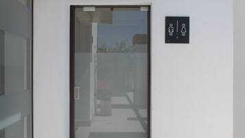 Entrada de um edifício moderno com porta de vidro, placa indicando sanitários masculino e feminino, e piso revestido de azulejos.