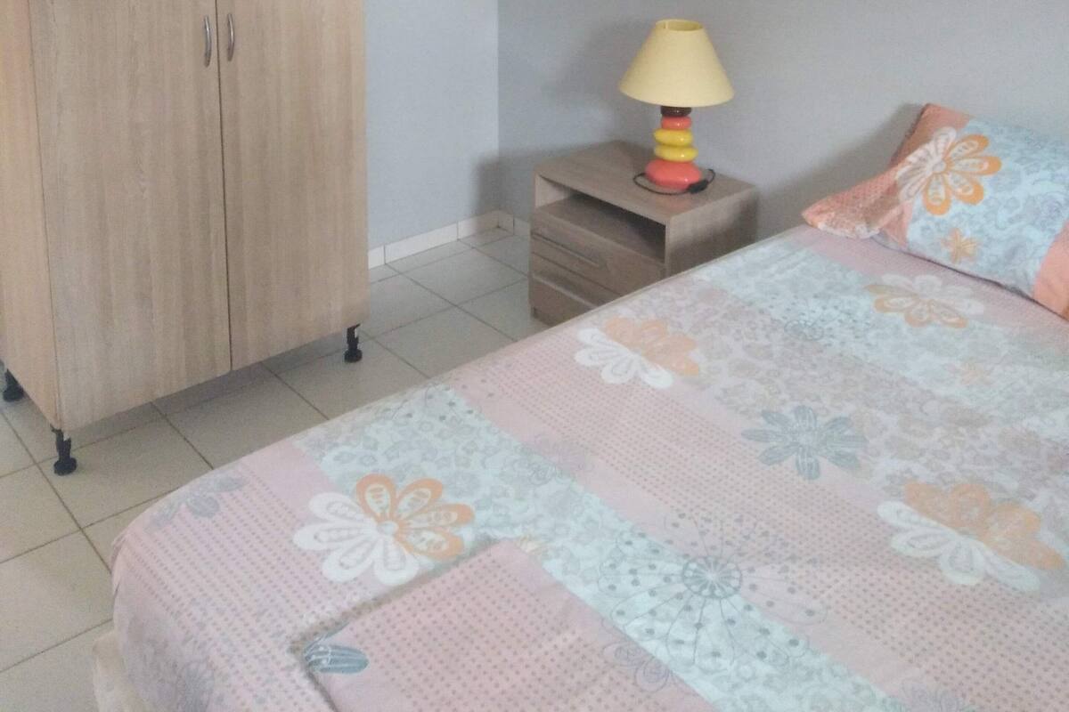 Quarto com cama, mesa de cabeceira com abajur, guarda-roupa e colcha estampada.