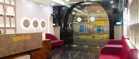 Uma área de recepção com balcão de mármore identificado como “RECEPTION”, assentos vermelhos e parede de vidro com vista para um edifício.
