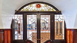 Conjunto de portas duplas com padrões de vidro elaborados e moldura de madeira, dando acesso a um espaço interior com piso ladrilhado.