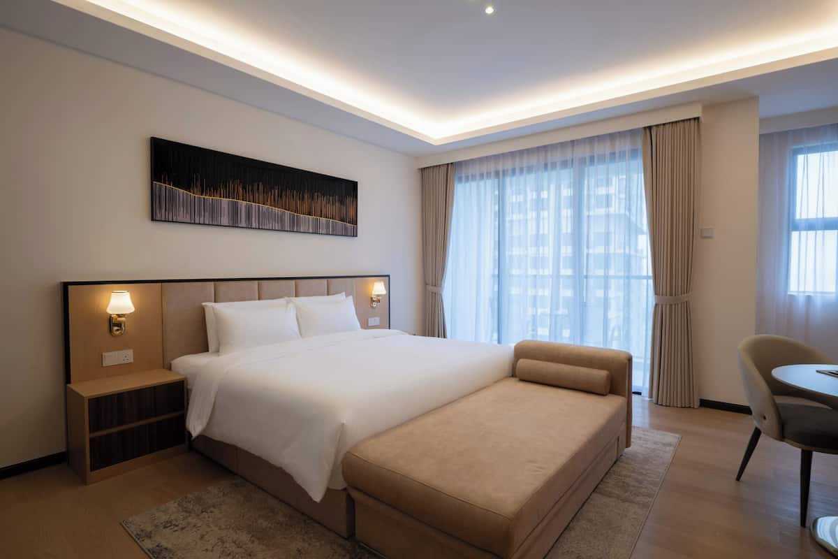 Quarto de hotel moderno com uma cama grande, abajures, uma pequena mesa e vista da cidade através de cortinas leves.