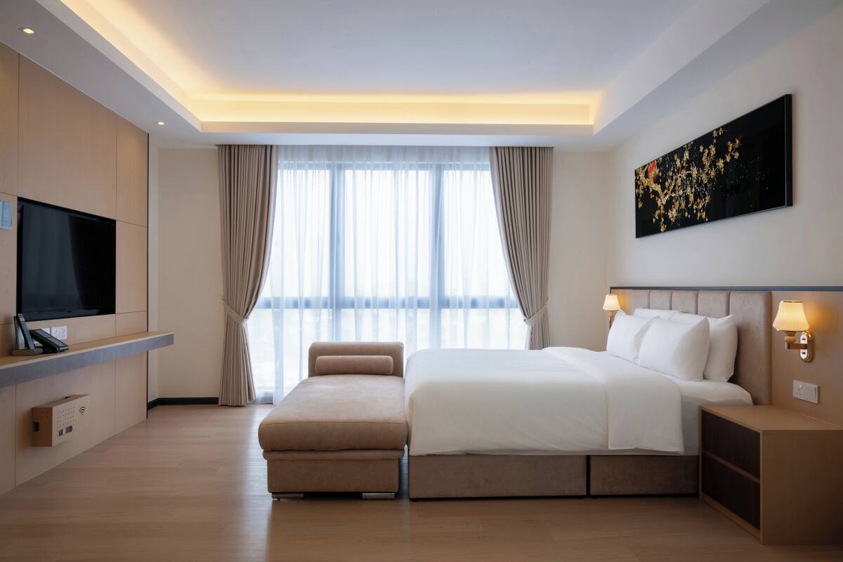 Quarto de hotel com cama, luminárias de cabeceira, televisão de tela plana, um pequeno espaço para sentar e uma grande janela com cortinas.