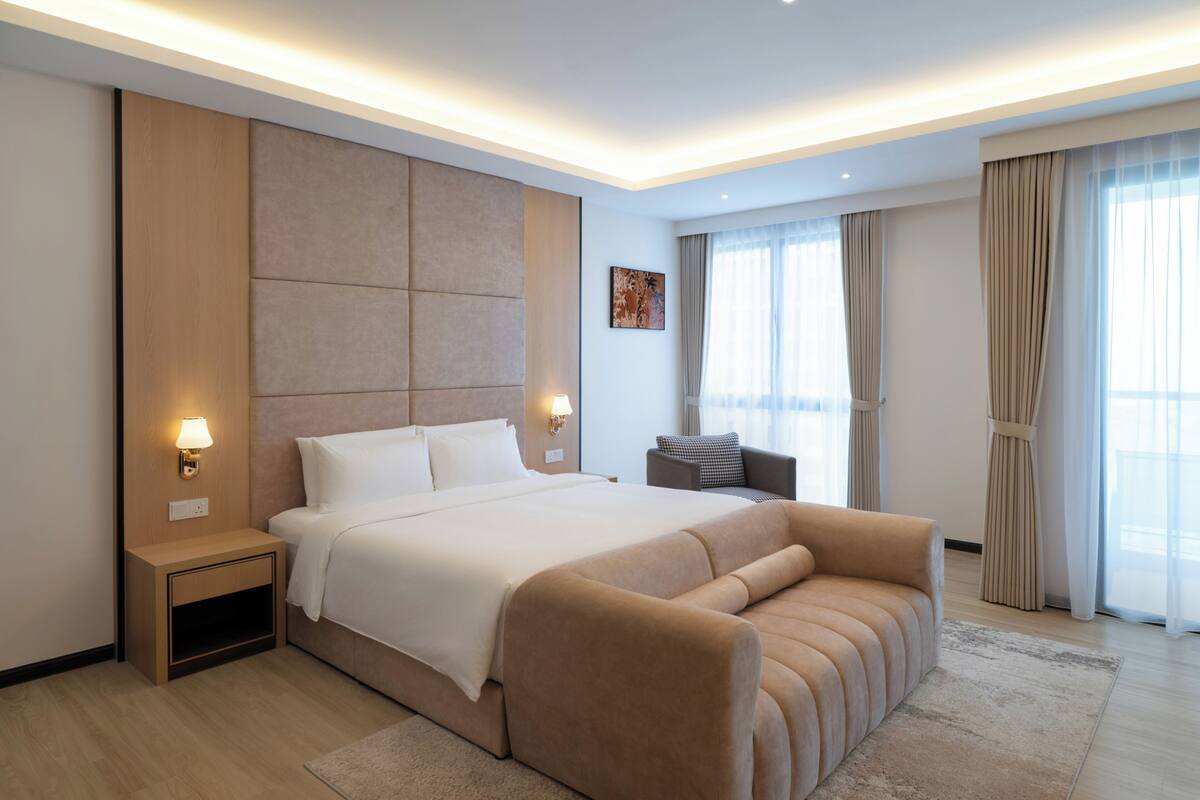 Quarto de hotel moderno com uma cama grande, um sofá, uma mesa de cabeceira e uma luminária na parede.