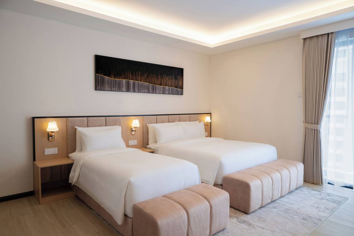 Quarto de hotel com duas camas, um banco, uma obra de arte na parede e uma mesa de cabeceira com luminária.