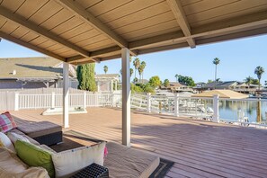 Um terraço coberto ao ar livre com piso de madeira, grades brancas e vista para um porto com barcos e palmeiras.