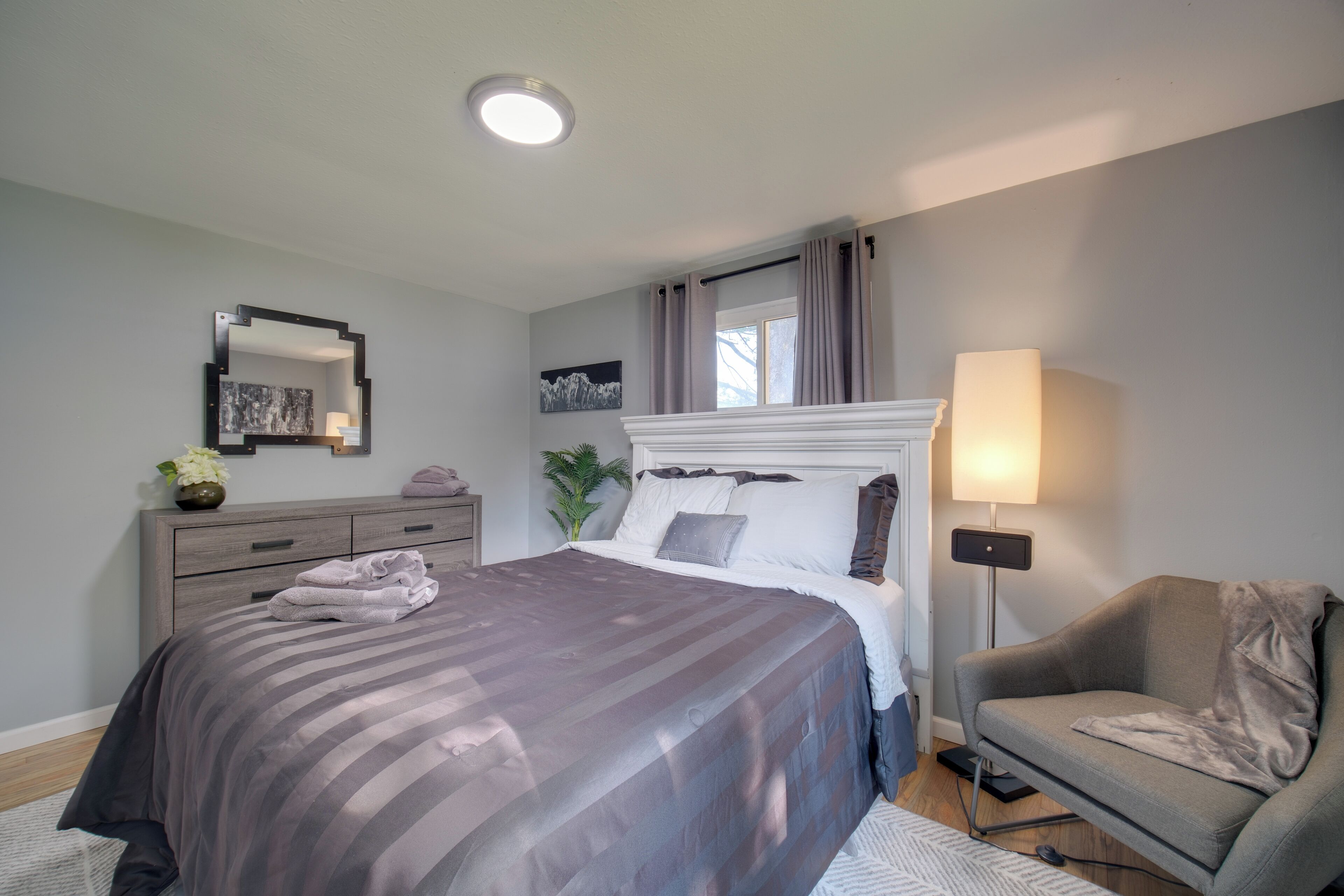 Quarto com cama, uma cadeira, mesa de cabeceira, luminária e janela com cortinas.