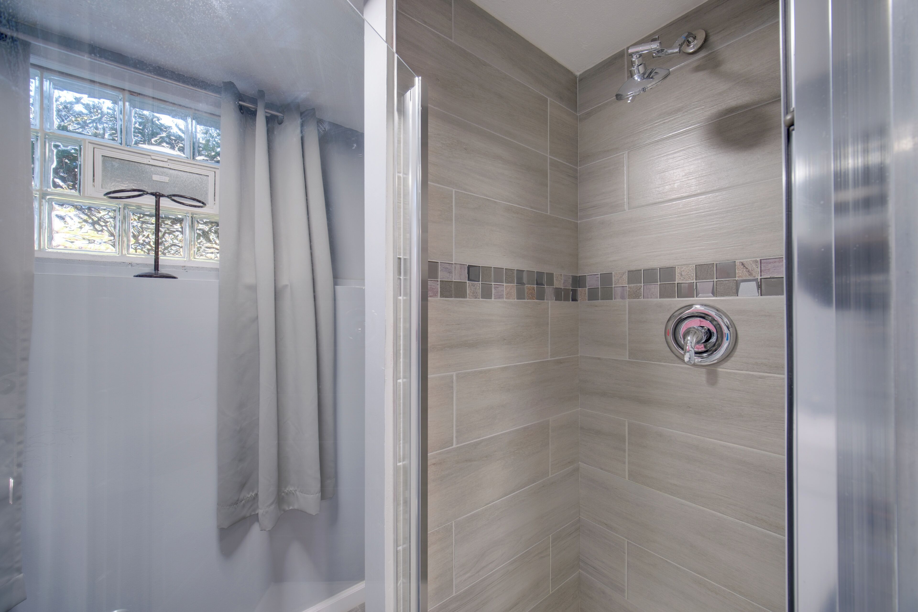 Chuveiro com porta de vidro, janela com cortinas e painel de azulejos.