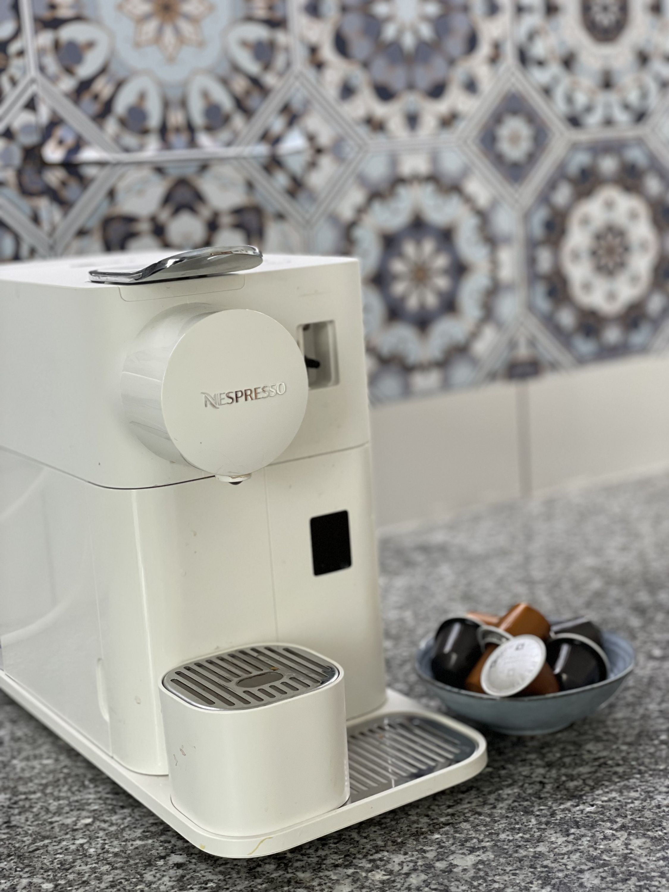 Máquina de café Nespresso branca sobre bancada de cozinha com painel de azulejos decorativo.