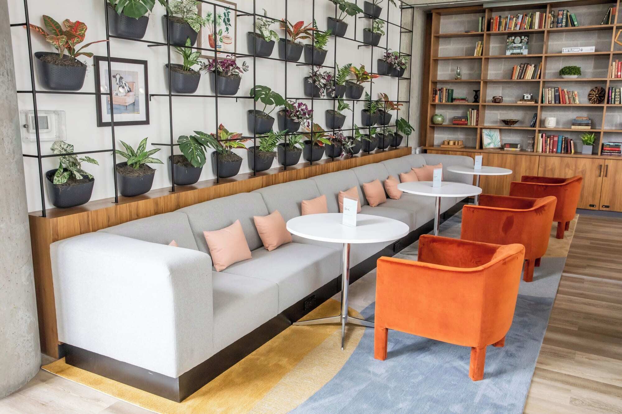 Uma área de estar moderna com um sofá cinzento, cadeiras laranja e uma estante repleta de livros e objetos decorativos.
