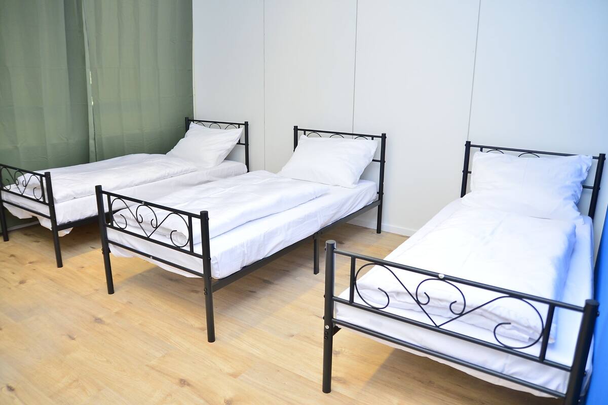 Três camas de solteiro com estruturas de metal e roupa de cama branca num quarto com soalho de madeira e cortina verde.