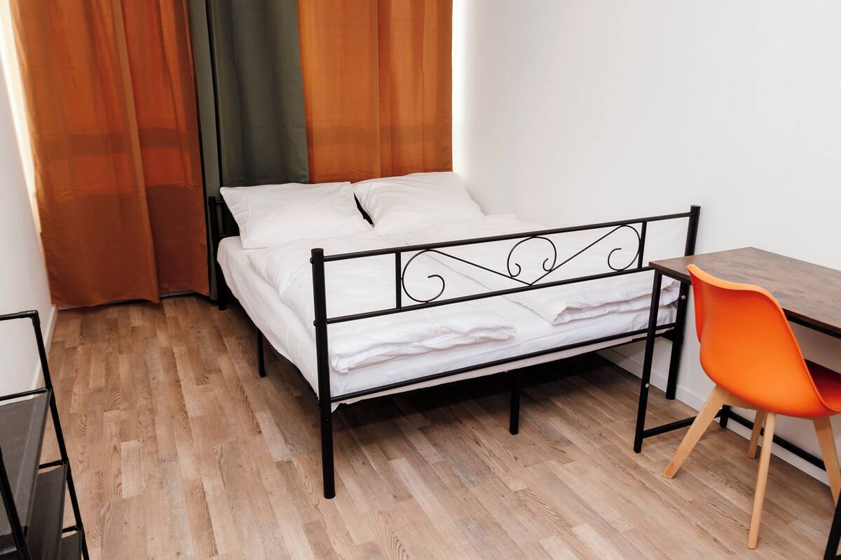 Uma cama de solteiro com estrutura de metal, uma escrivaninha de madeira com uma cadeira e uma janela com cortinas laranja.