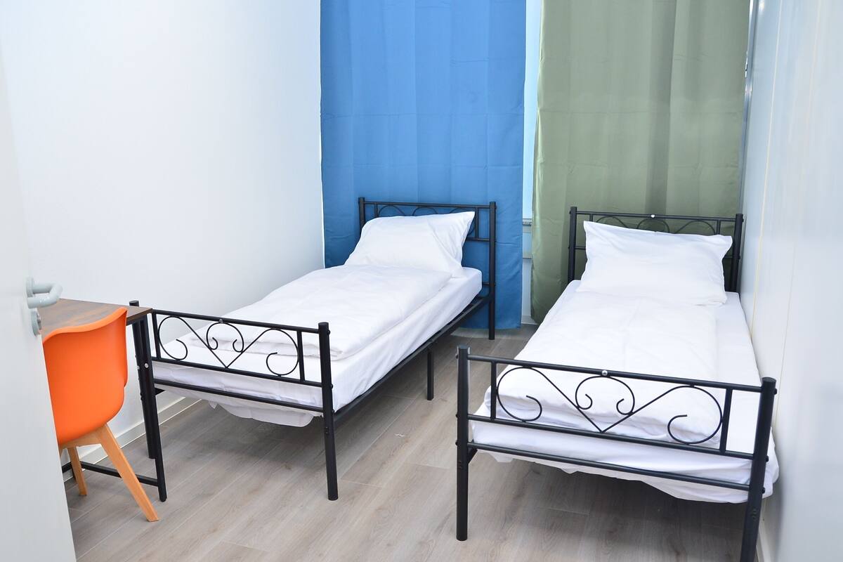 Duas camas de solteiro com estrutura de metal e roupa de cama branca, uma cadeira laranja pequena e uma escrivaninha de madeira num quarto com soalho de madeira claro.
