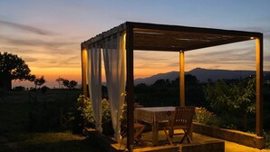 Um gazebo de madeira com uma mesa e cadeiras, situado sobre um caminho de cascalho, com vista para montanhas distantes sob um céu crepuscular.