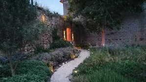 Um caminho de cascalho serpenteia por um jardim, com um muro de tijolos e um muro de pedra de um lado, e diversas plantas e arbustos do outro.