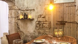 Uma mesa de jantar rústica posta com pratos, copos e uma cesta de pão, com uma parede de pedra e porta de madeira ao fundo.