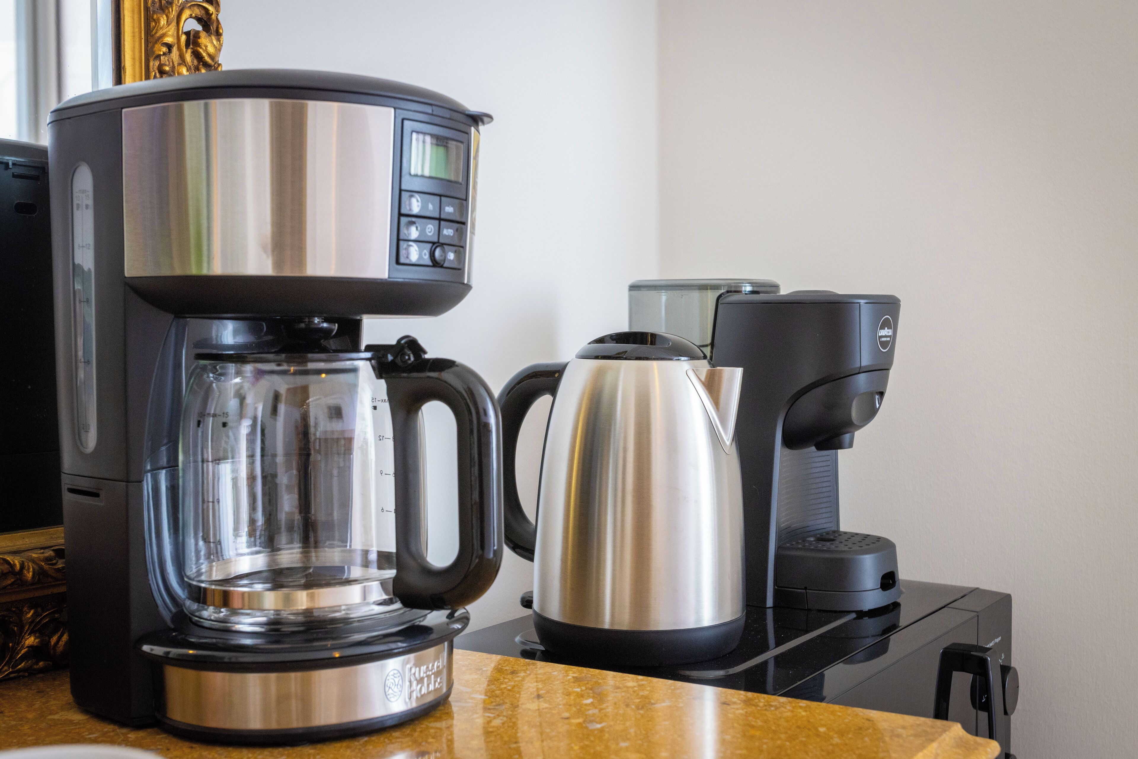Uma cafeteira com visor digital, uma chaleira de aço inoxidável e uma jarra de café em uma bancada de cozinha.