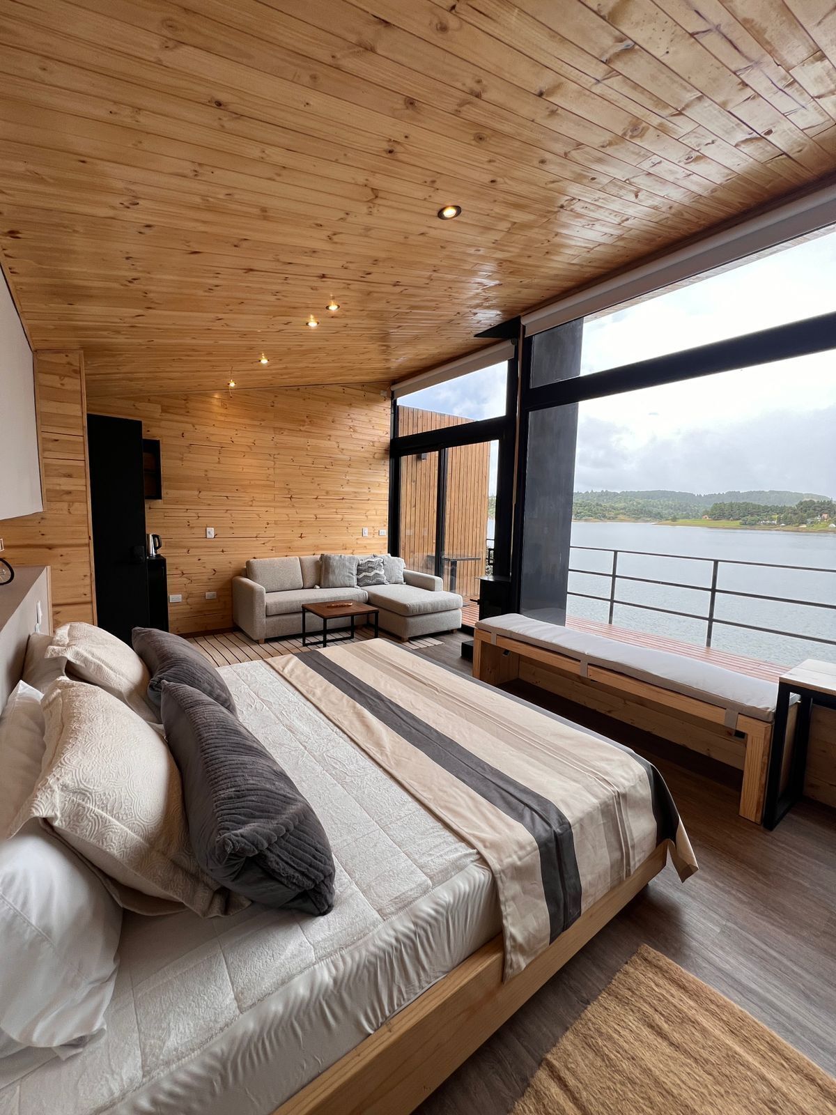 Quarto moderno com uma cama grande, um sofá e um banco perto de uma janela com vista para a água.