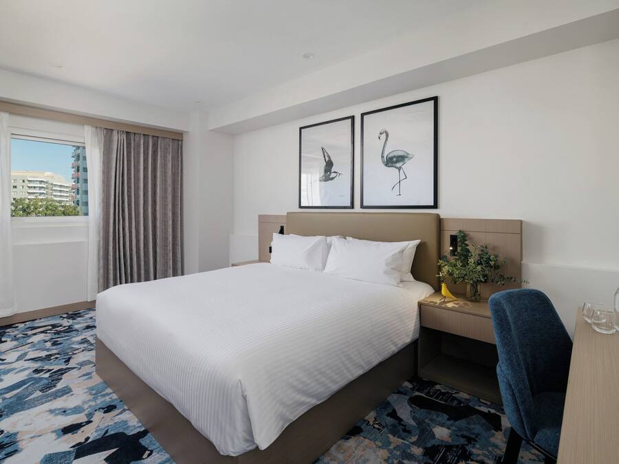 Quarto de hotel com uma cama grande, uma escrivaninha com cadeira, uma janela com cortinas e duas obras de arte penduradas na parede.