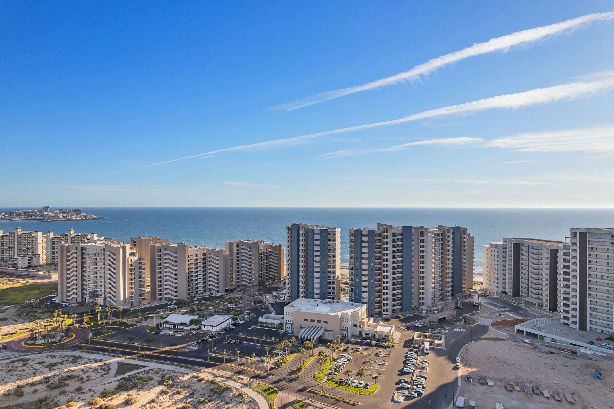 Zona residencial costeira com edifícios altos, um parque de estacionamento e uma praia ao fundo.
