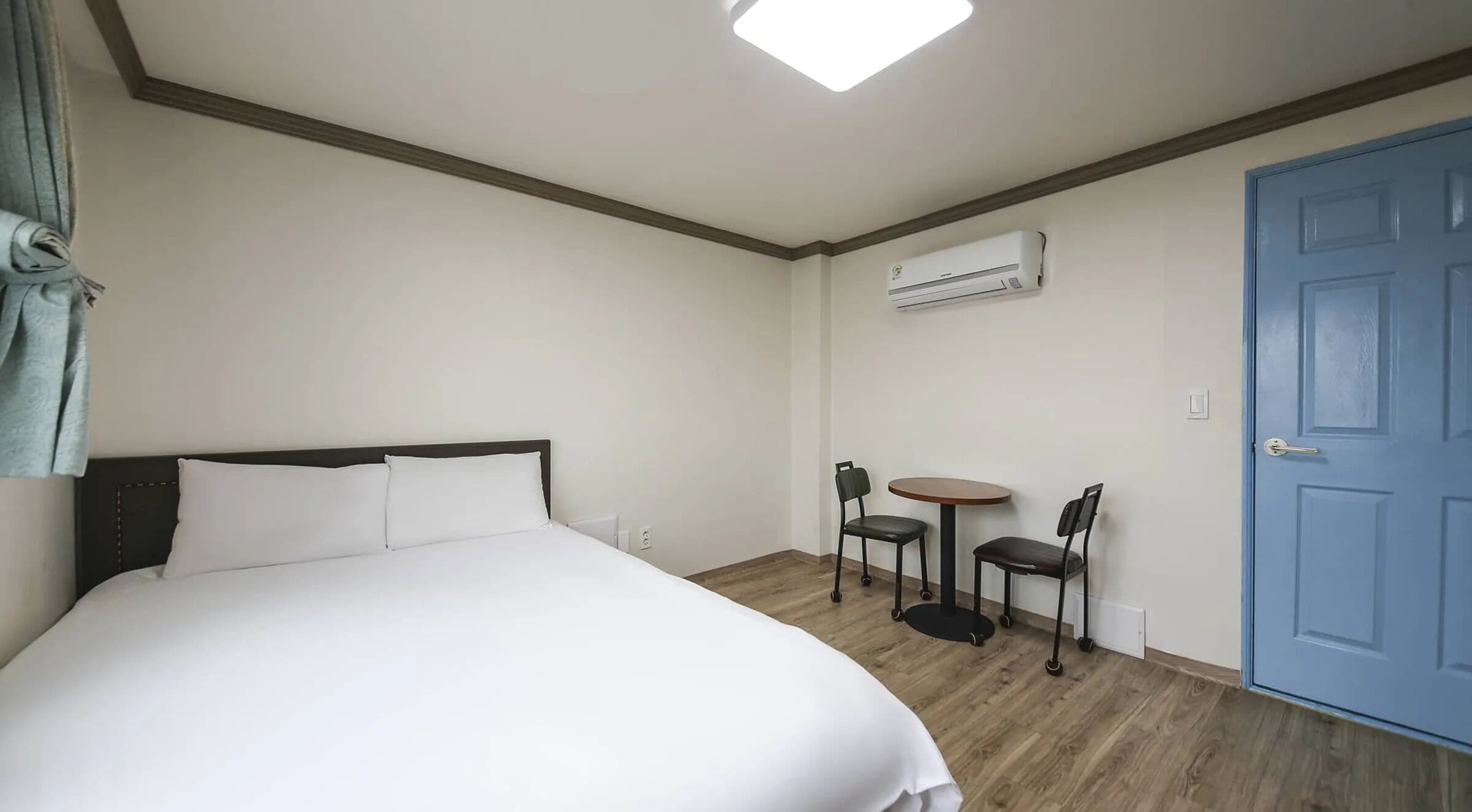 Quarto de hotel com cama, uma mesinha com duas cadeiras, um aparelho de ar condicionado e uma porta azul.