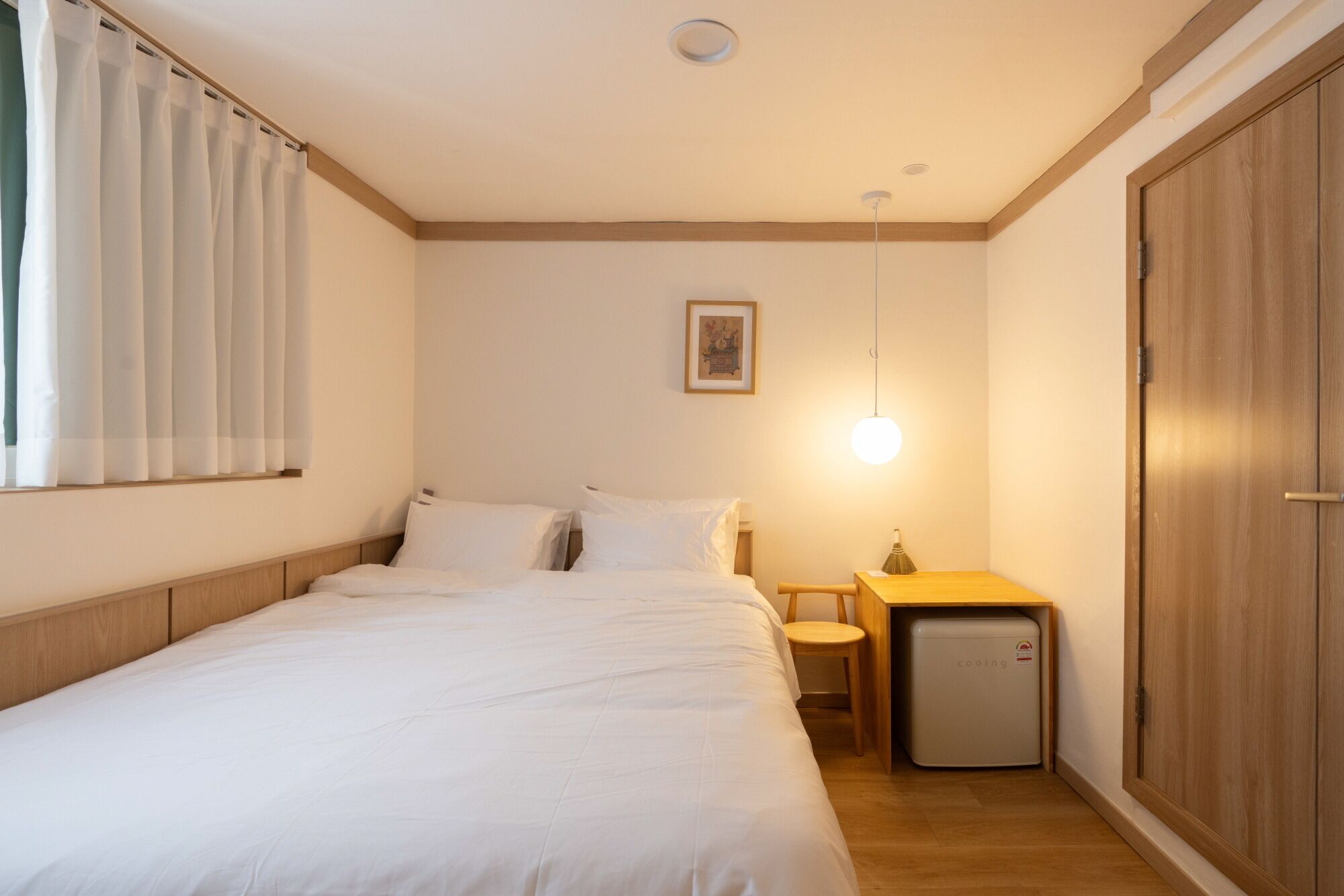 Quarto de hotel com cama, secretária pequena, cadeira, frigorífico e guarda-roupa.
