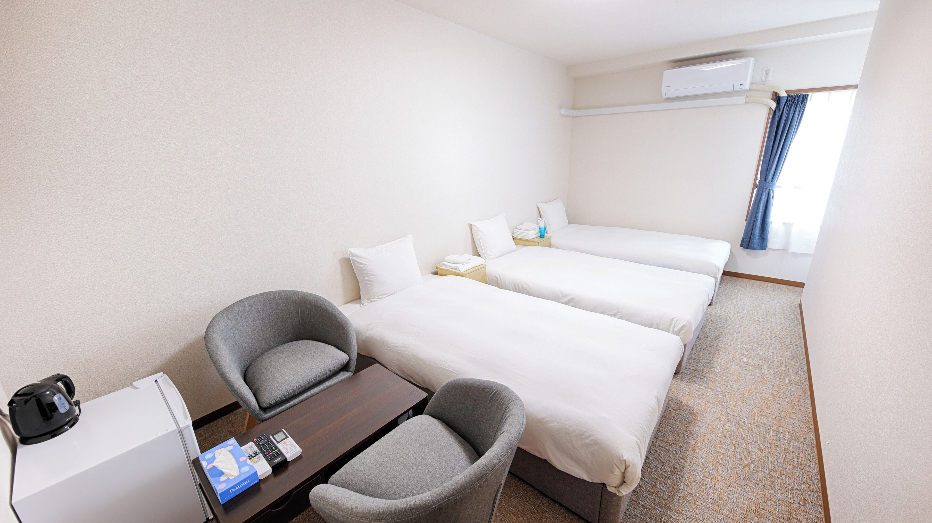 Quarto de hotel com uma escrivaninha, duas poltronas cinzentas, duas camas de solteiro, uma mesinha com uma cafeteira e uma caixa, e uma janela com cortinas.