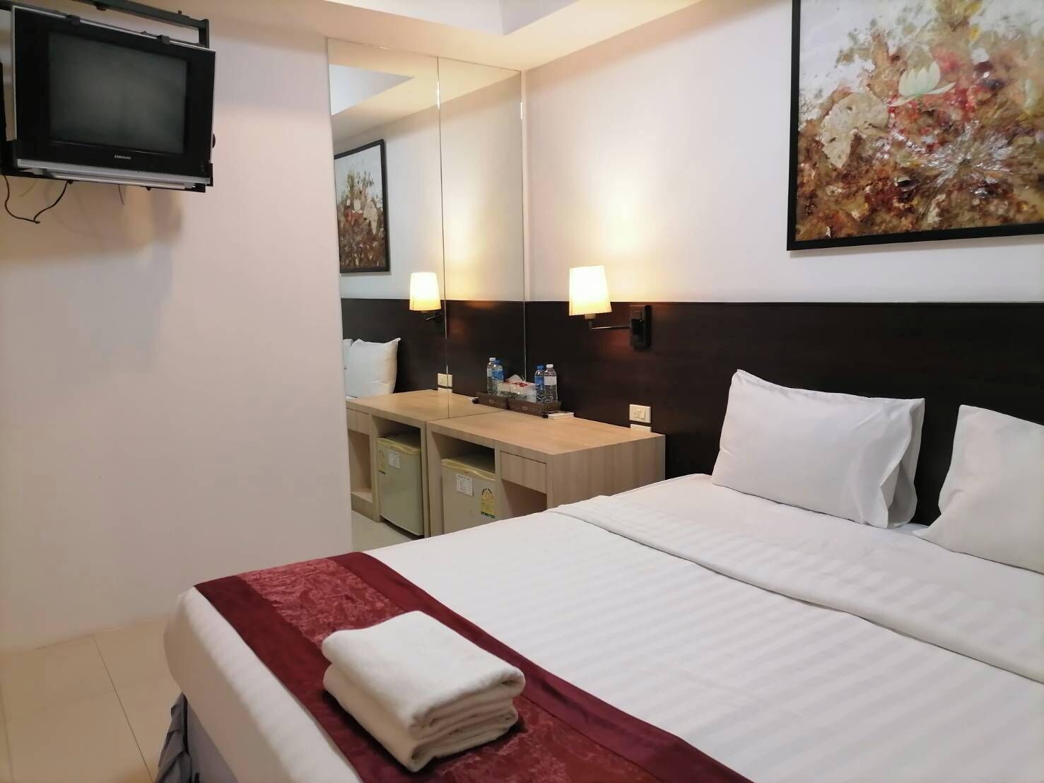 Quarto de hotel com cama, escrivaninha, espelho, televisão e duas luminárias fixadas na parede.