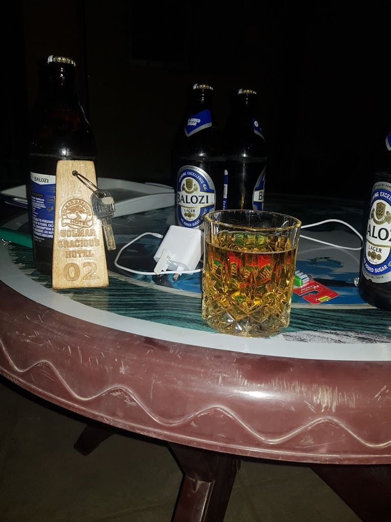 Uma mesa com duas garrafas de cerveja Balozi, um chaveiro de madeira, um copo com bebida e um objeto branco.